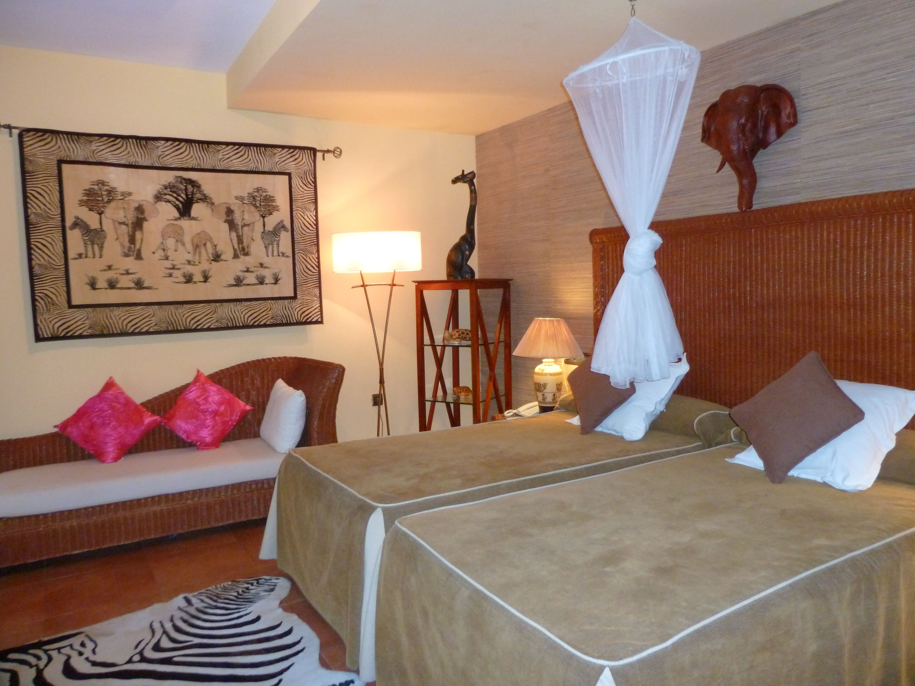 Quarto de hotel com duas camas, um sofá, um tapete com tema de zebra, um quadro de zebra fixado na parede, um abajur e uma cabeça de elefante decorativa.