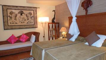 Quarto de hotel com duas camas, um sofá, um tapete com tema de zebra, um quadro de zebra fixado na parede, um abajur e uma cabeça de elefante decorativa.