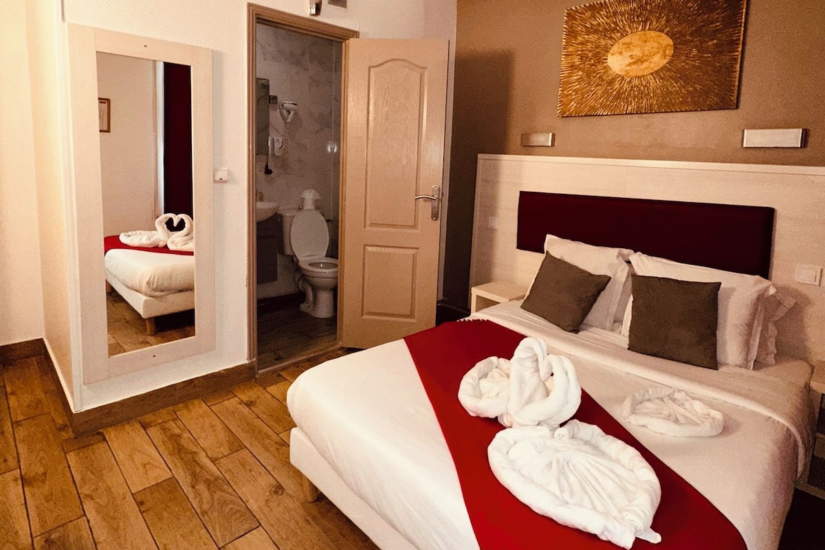 Quarto de hotel com uma cama grande, um cobertor vermelho, duas toalhas brancas, piso de madeira, banheiro com vaso sanitário e pia, e um espelho.