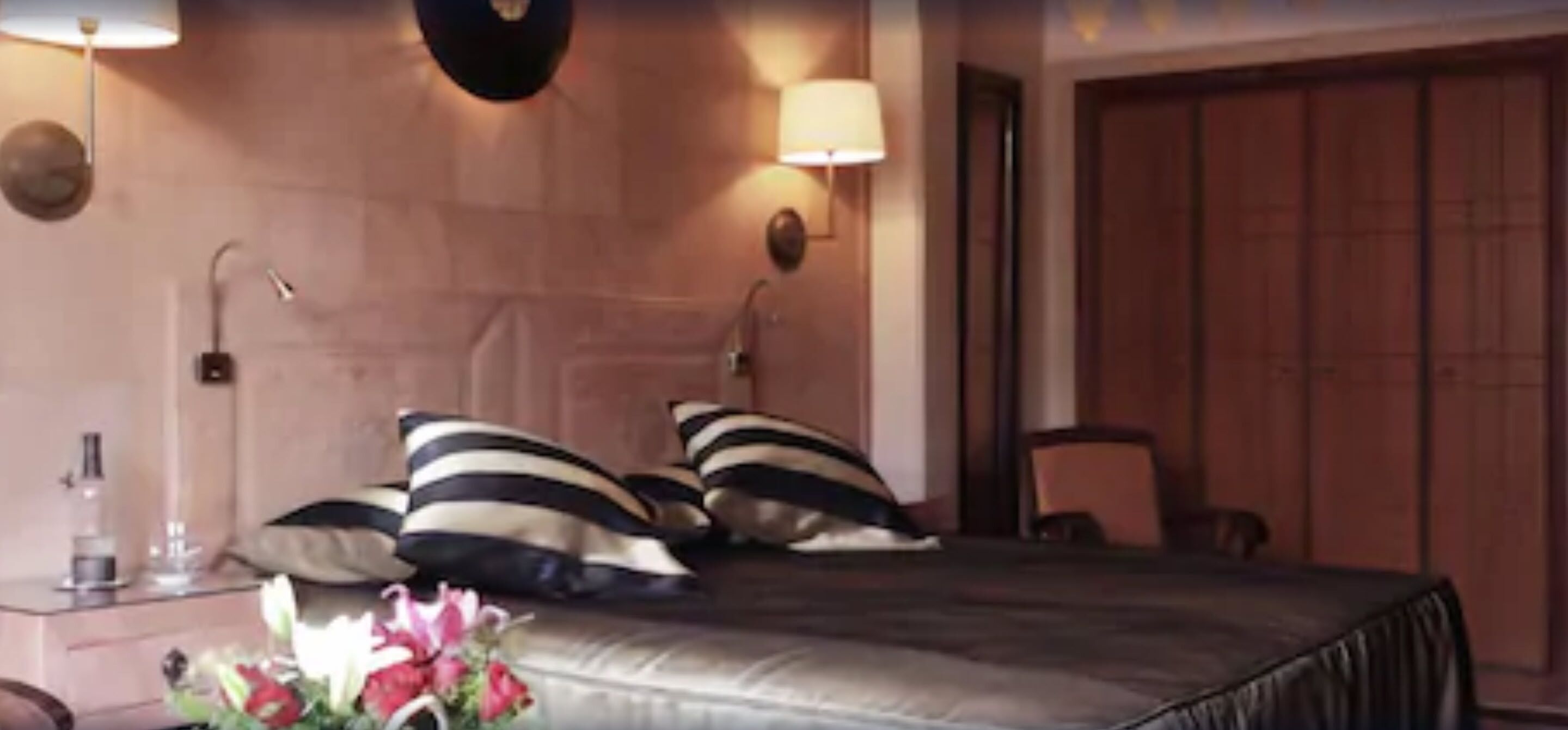 Quarto de hotel com uma cama grande, almofadas listradas, mesa de cabeceira com abajur e um vaso de flores.