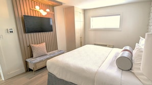 Quarto moderno com cama, um banco, televisão e janela.