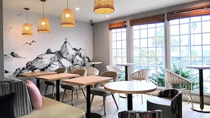Um café moderno com luminárias pendentes de vime, um mural de montanhas e janelas amplas com vista para palmeiras.