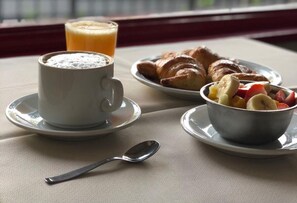 Uma mesa posta para o café da manhã, com uma xícara de café, croissants, uma tigela de frutas e um copo de suco de laranja.