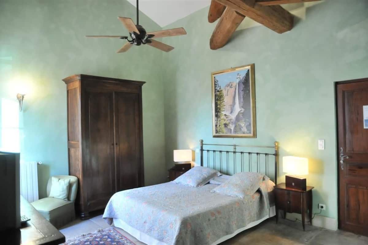 Um quarto com cama, mesa de cabeceira, cadeira, guarda-roupa, ventilador de teto e um quadro na parede.