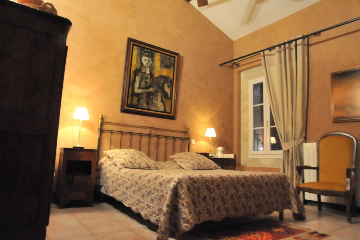 Quarto com uma cama, duas luminárias de cabeceira, uma cadeira, uma janela com cortinas e um quadro na parede.