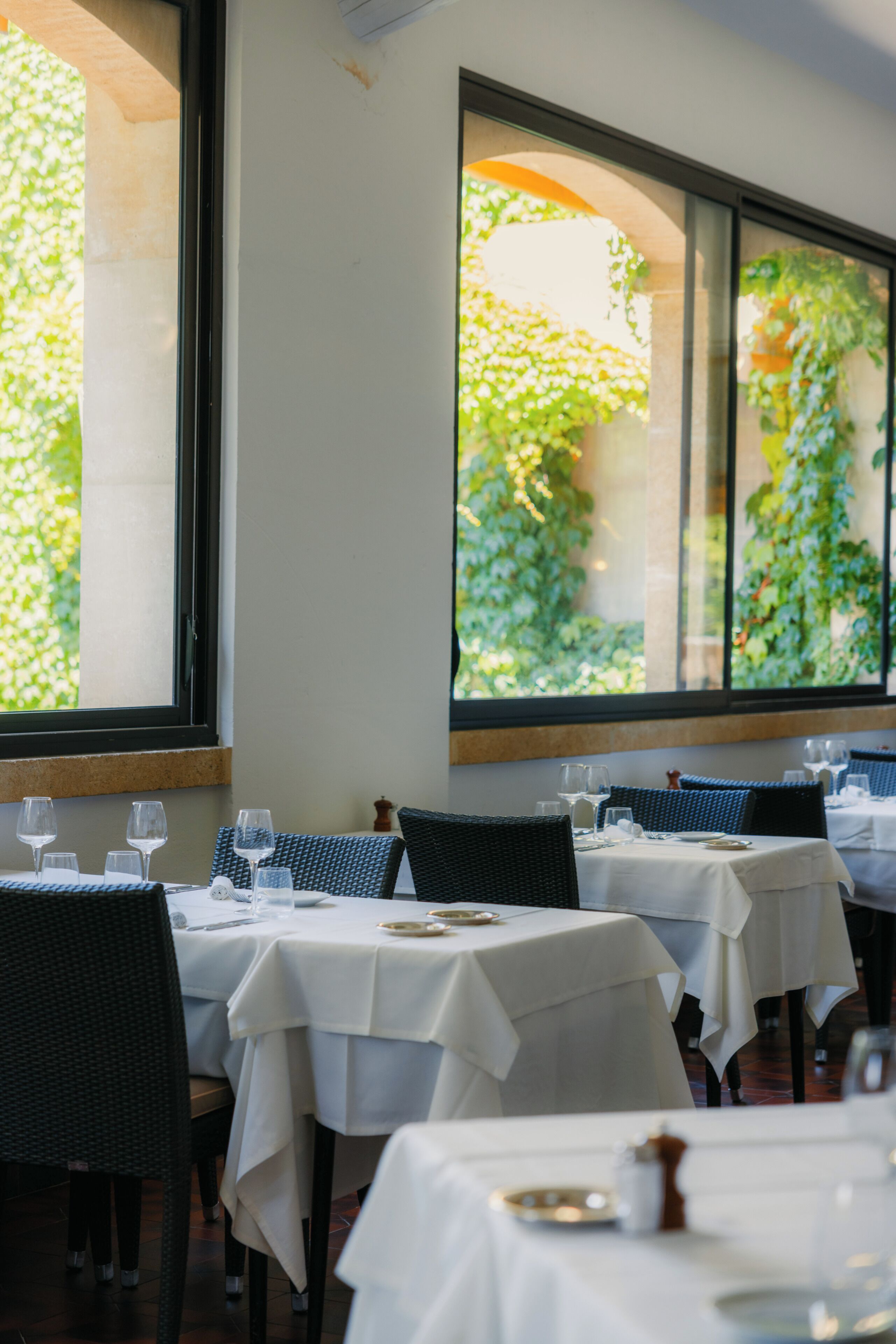 Interior de restaurante com mesas preparadas para refeições, grandes janelas com vegetação no exterior e um design moderno e elegante.