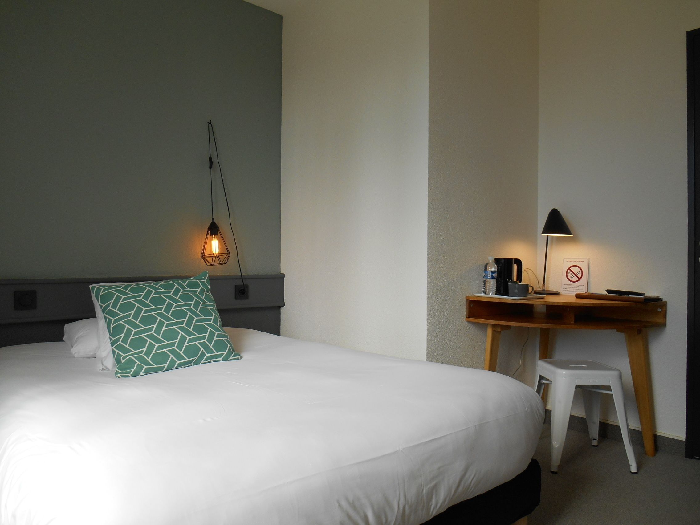 Quarto de hotel com cama, escrivaninha, cadeira e mesa de cabeceira.