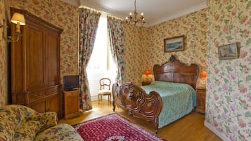 Quarto com papel de parede floral, guarda-roupa de madeira, cama com cabeceira, tapete vermelho e televisão sobre um móvel de madeira.
