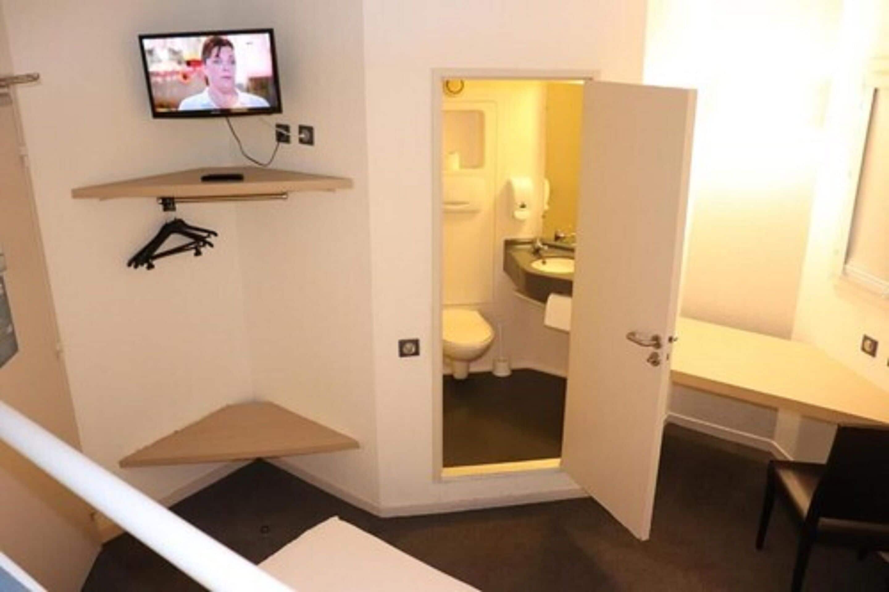 Quarto pequeno e compacto, com televisão na parede, uma mesinha e uma porta de acesso a um banheiro.