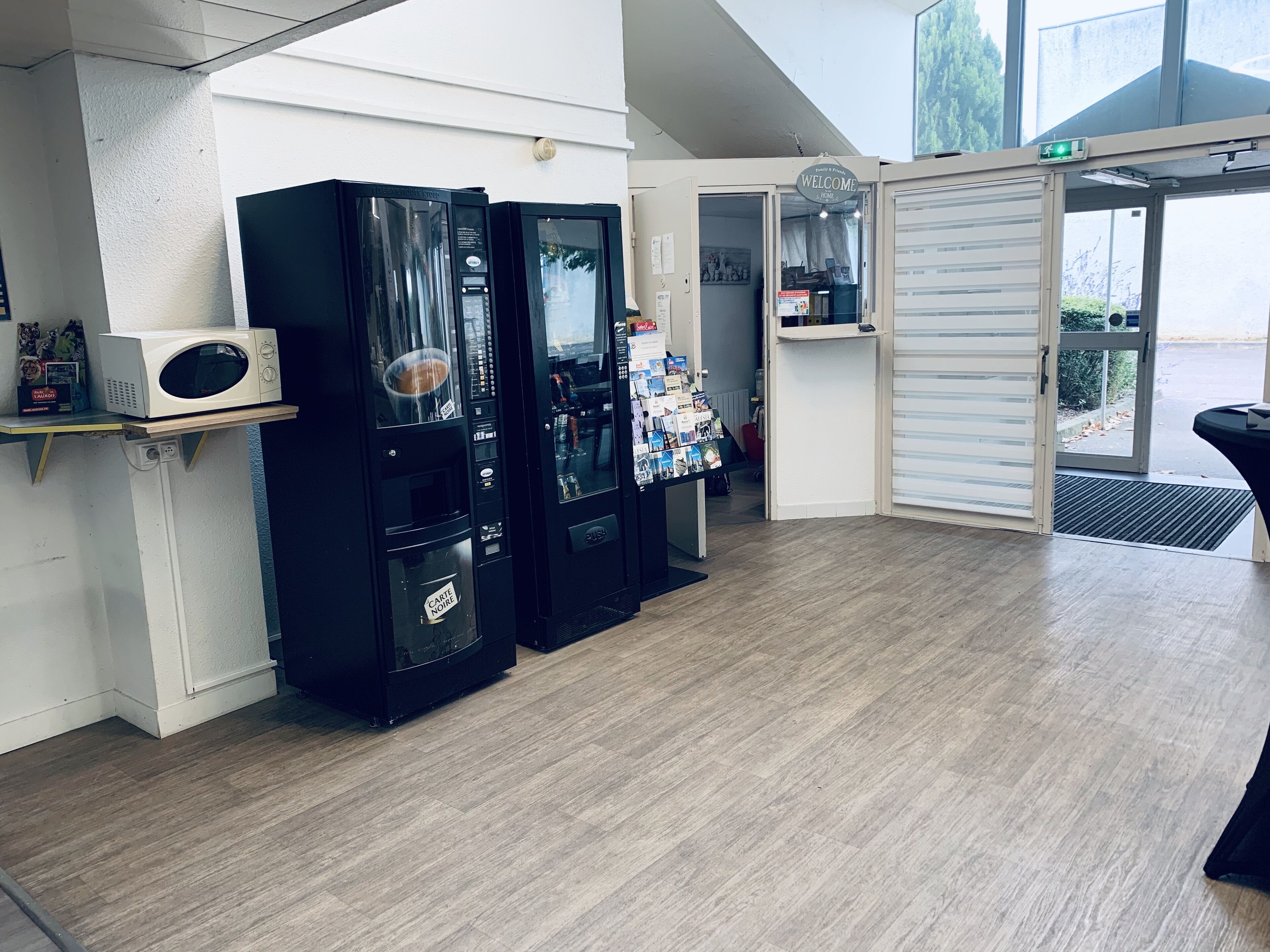 Quarto com piso de madeira, máquina de vendas automática, micro-ondas e porta com placa de 'Welcome'.