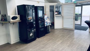 Quarto com piso de madeira, máquina de vendas automática, micro-ondas e porta com placa de 'Welcome'.