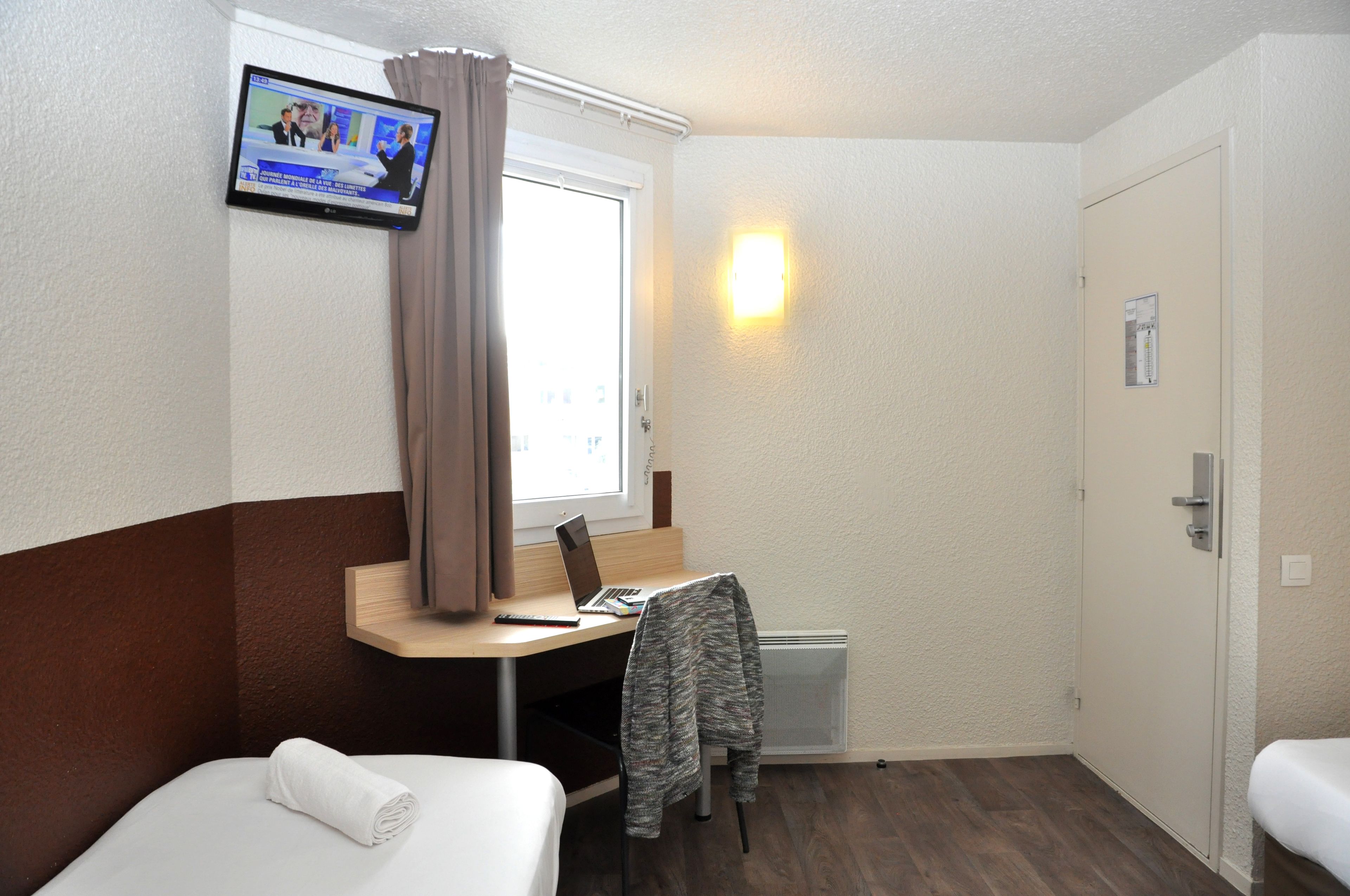 Quarto de hotel com cama, escrivaninha com laptop, televisão na parede e janela com cortinas.