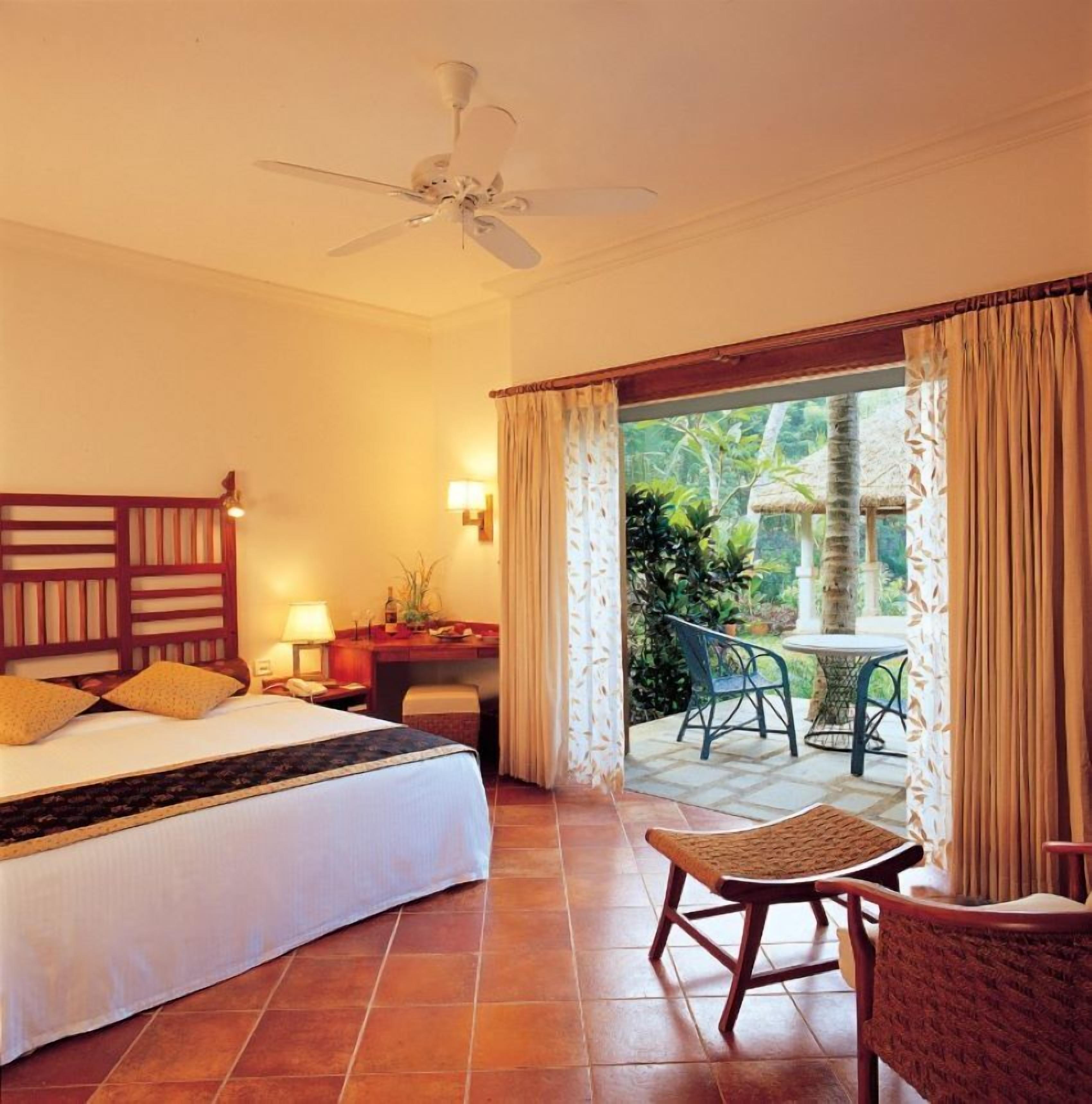 Quarto de hotel com uma cama, uma secretária com abajur, uma cadeira e vista para um jardim através de uma porta de vidro.