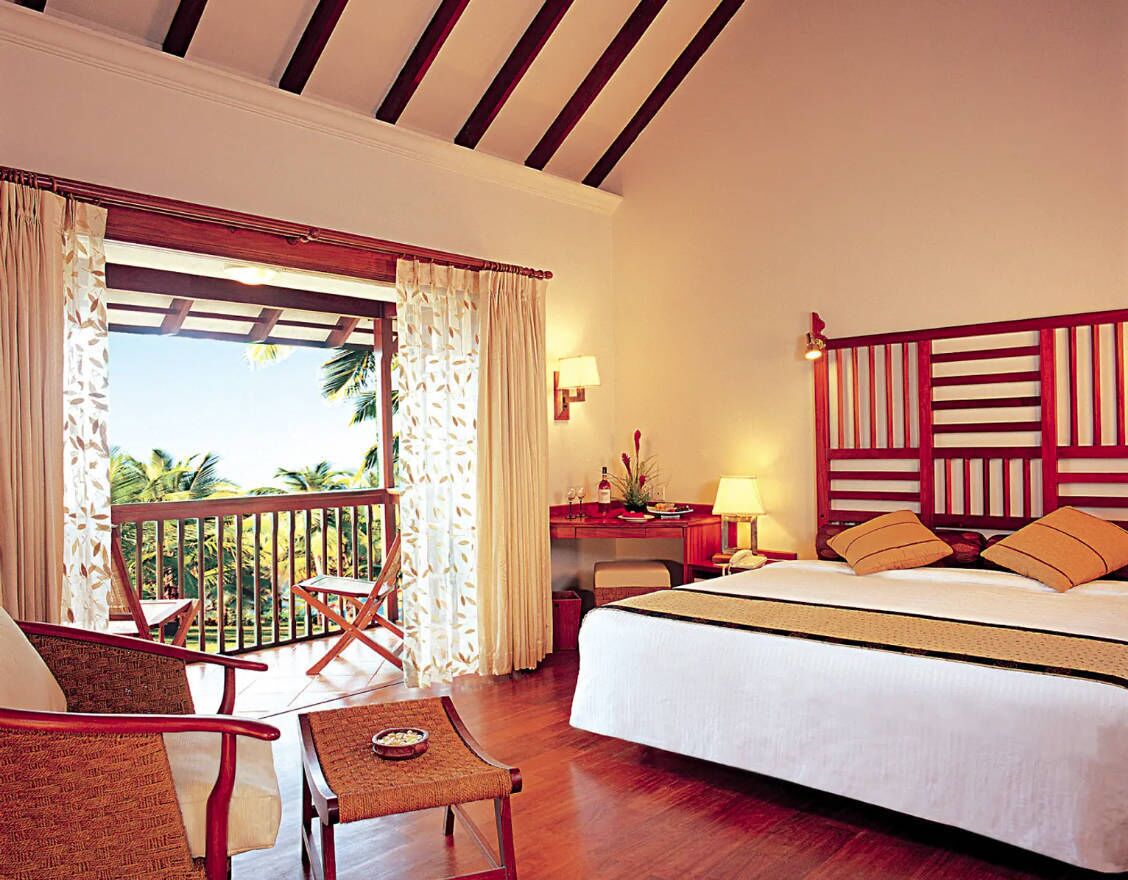 Quarto de hotel com cama, escrivaninha, cadeira, mesa pequena e varanda com vista para palmeiras.