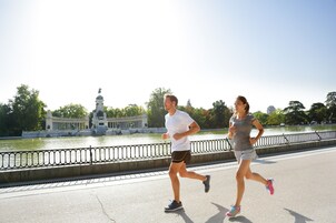 Duas pessoas correndo em uma ponte, com um corpo de água e uma estátua ao fundo.