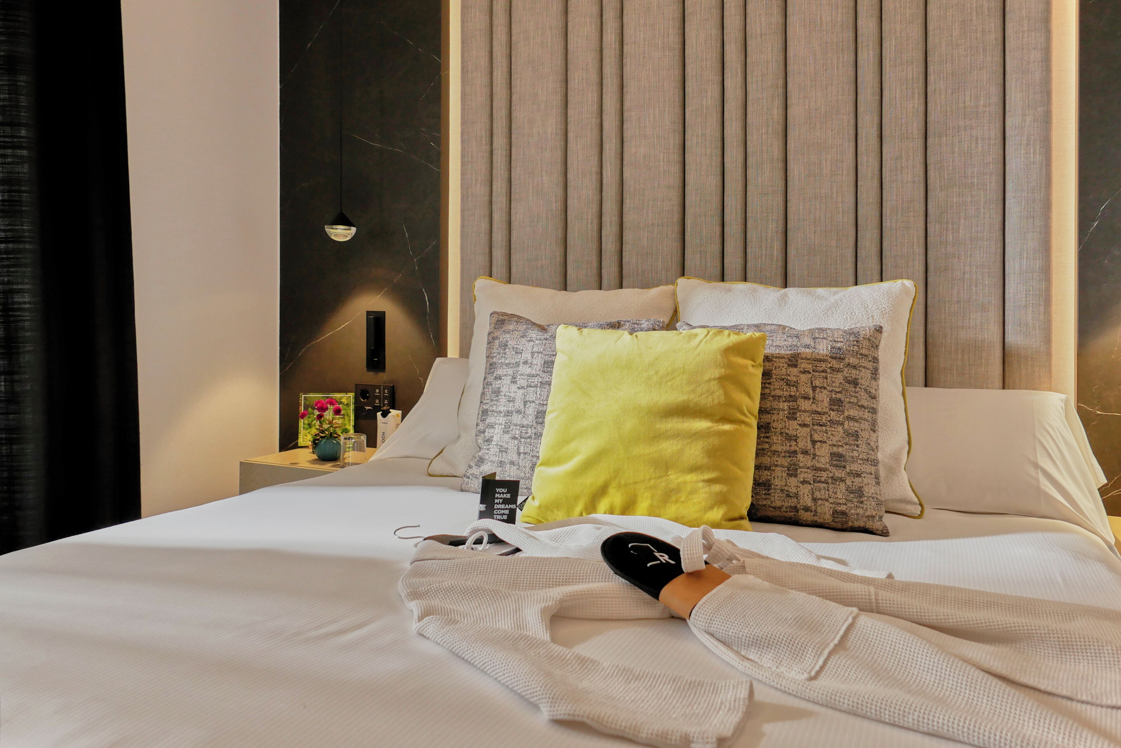 Cama bem arrumada com almofadas, mesa de cabeceira com um vaso de flores e luminária fixada na parede.
