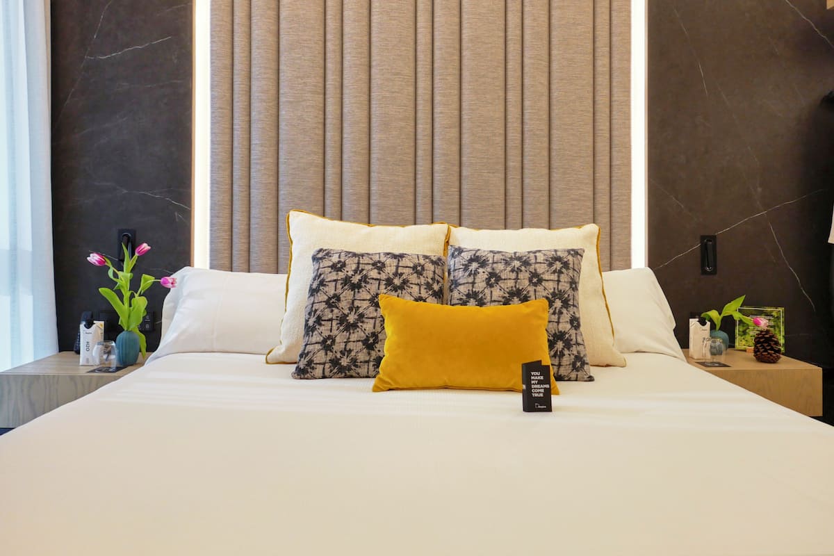 Cama bem arrumada com vários travesseiros, mesa de cabeceira com um vaso de flores e um criado-mudo com uma pequena planta.
