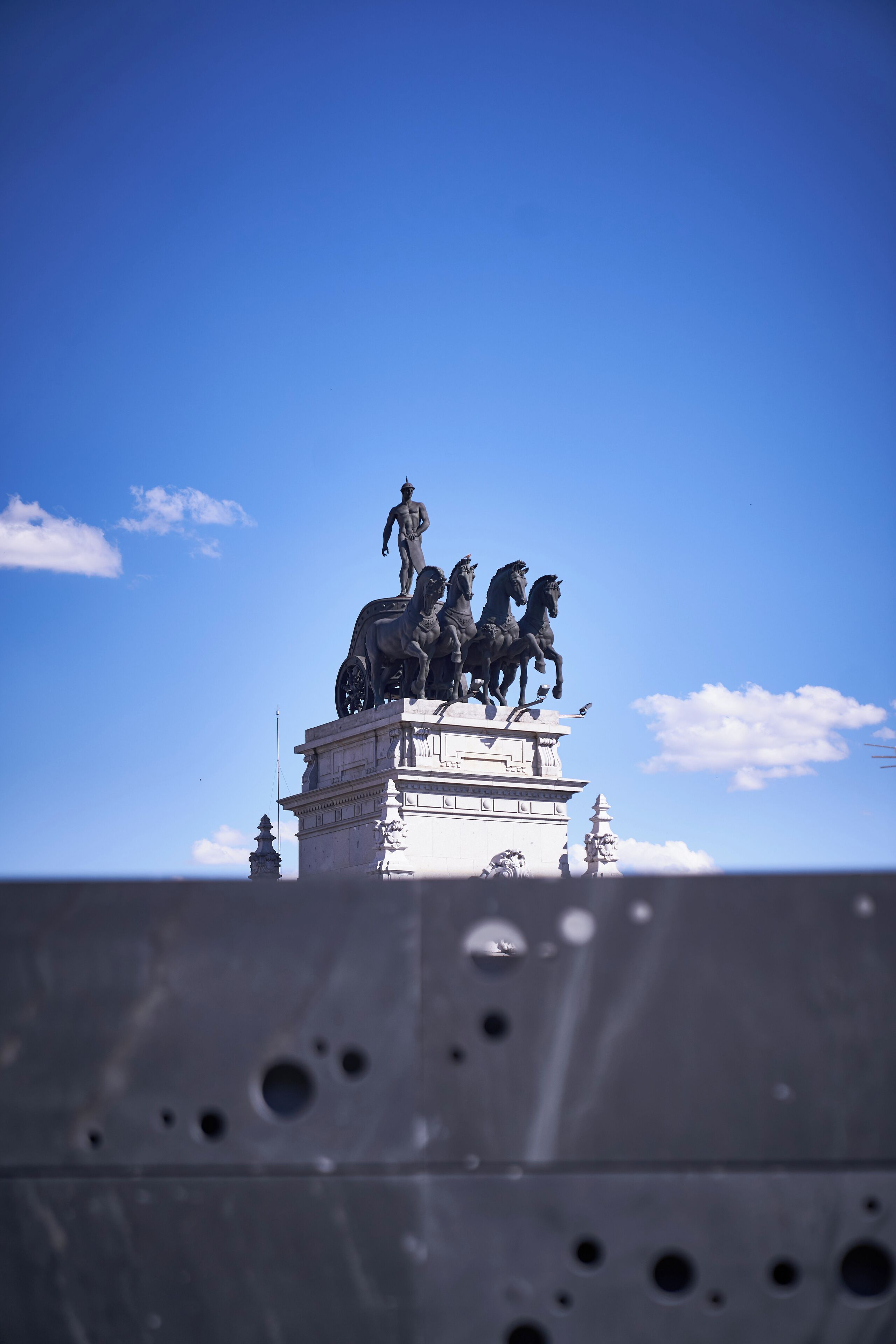 Estátua de um homem a cavalo com dois cavalos, assente num pedestal contra um céu azul límpido.