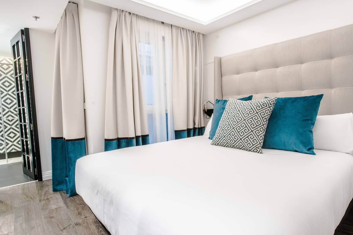 Quarto de hotel moderno com uma cama grande, uma mesa de cabeceira com abajur e vista para a cidade pela janela.