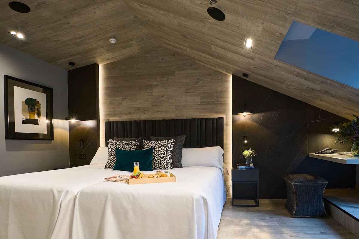 Quarto moderno com teto de madeira, cama com roupa de cama branca, mesa de cabeceira com um vaso de flores e um banquinho pequeno.
