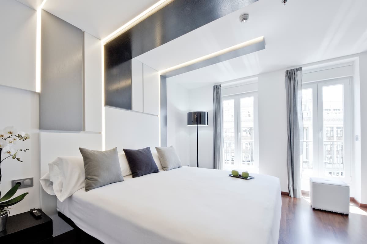 Quarto de hotel moderno com uma cama grande, mesa de cabeceira e vista da cidade através de amplas janelas.