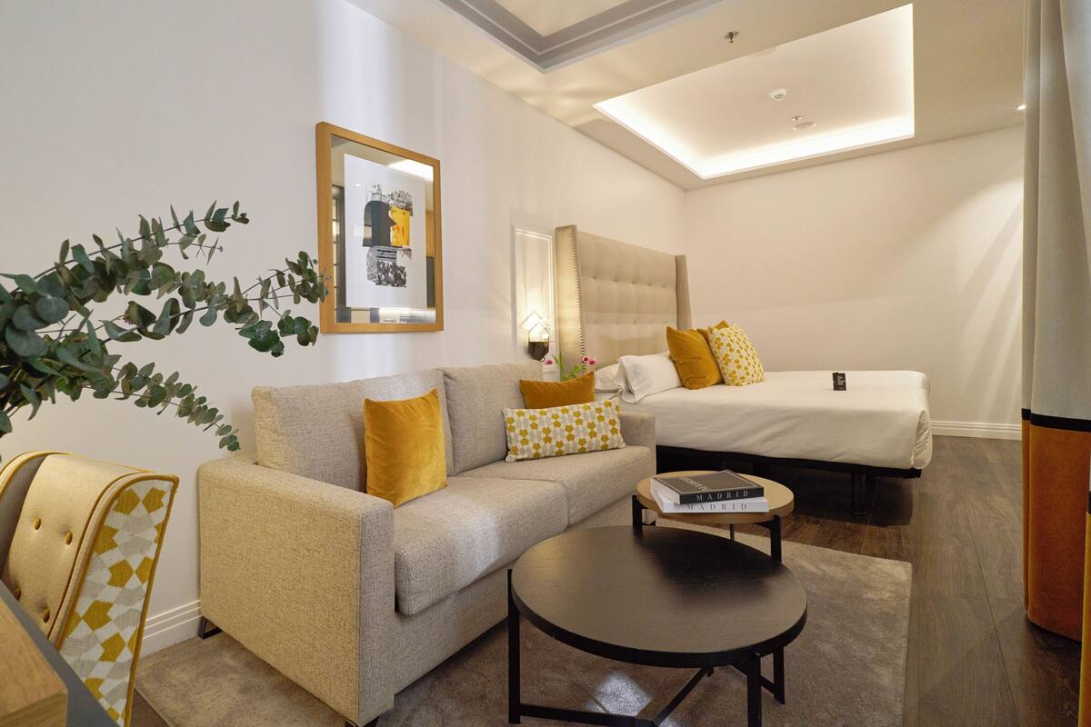 Quarto de hotel moderno com sofá, cama, mesa de centro redonda e um espelho grande.