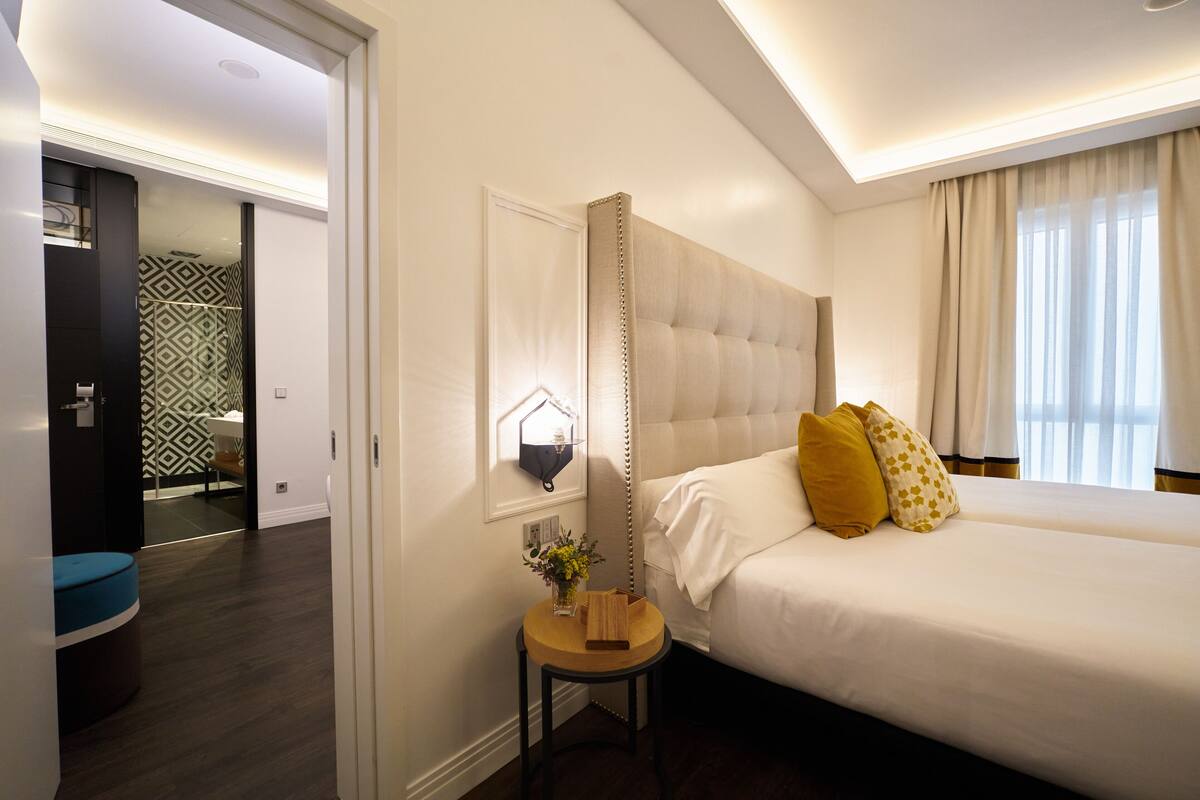 Quarto de hotel moderno com uma cama grande, uma mesinha de cabeceira com um vaso de flores e vista para o corredor.