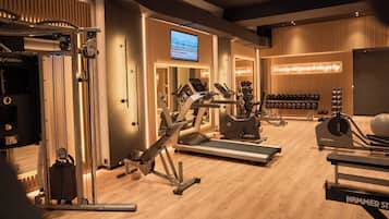 Academia com diversos equipamentos de ginástica, uma tela de televisão e arte motivacional nas paredes.