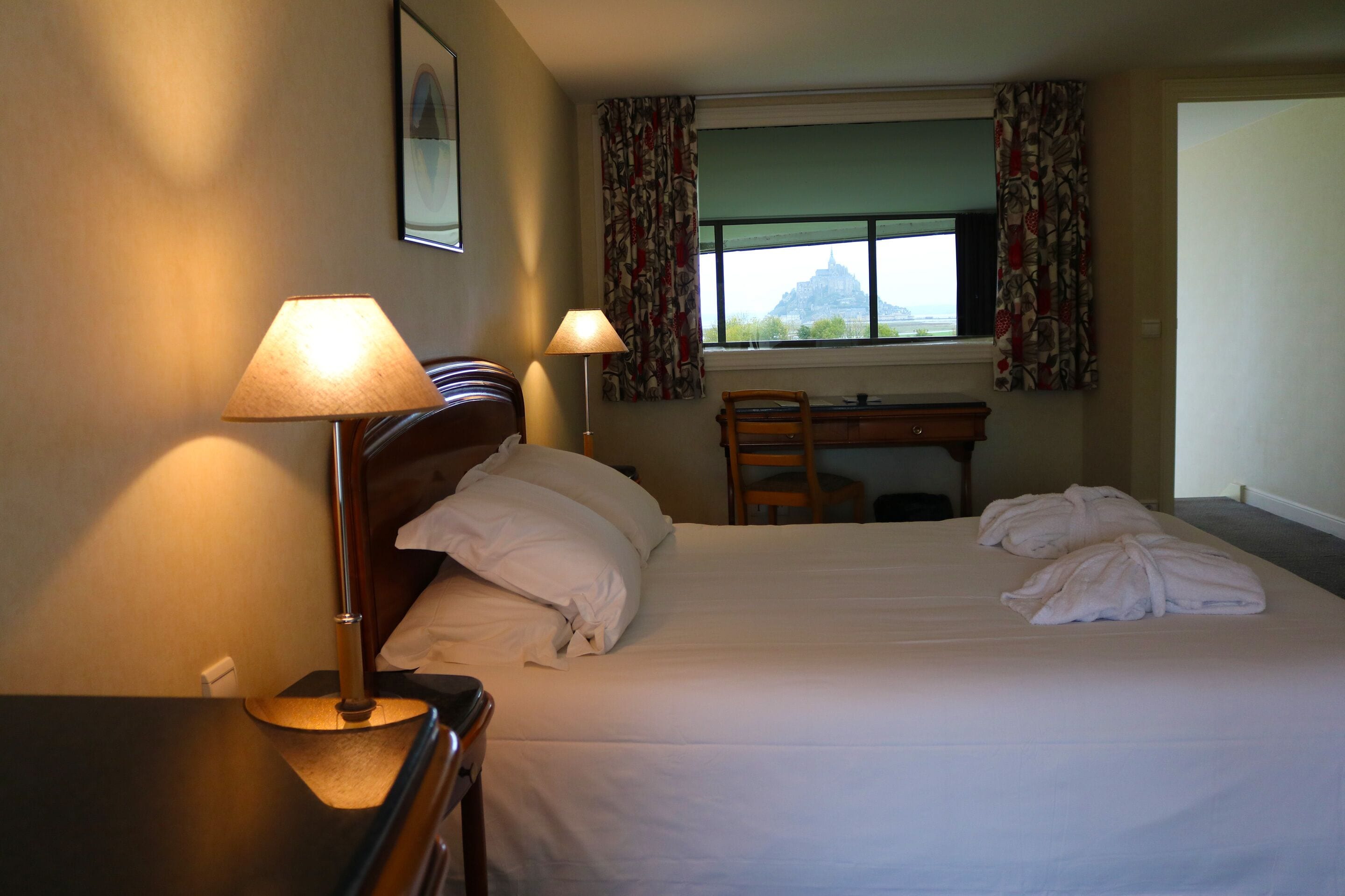 Quarto de hotel com cama, duas luminárias de cabeceira, escrivaninha, cadeira e janela com cortinas.