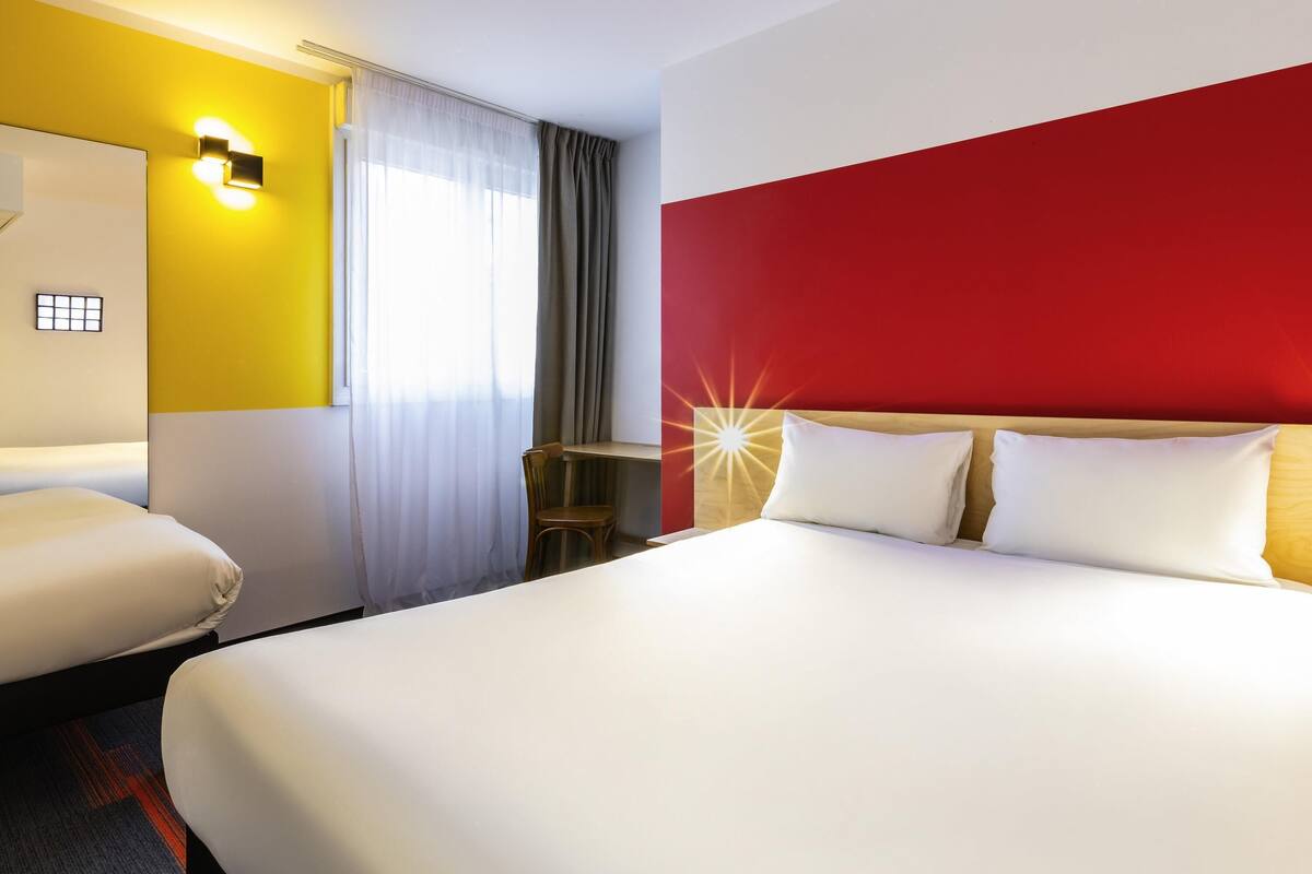 Quarto de hotel com uma cama grande, uma escrivaninha, uma cadeira e uma janela com cortinas.