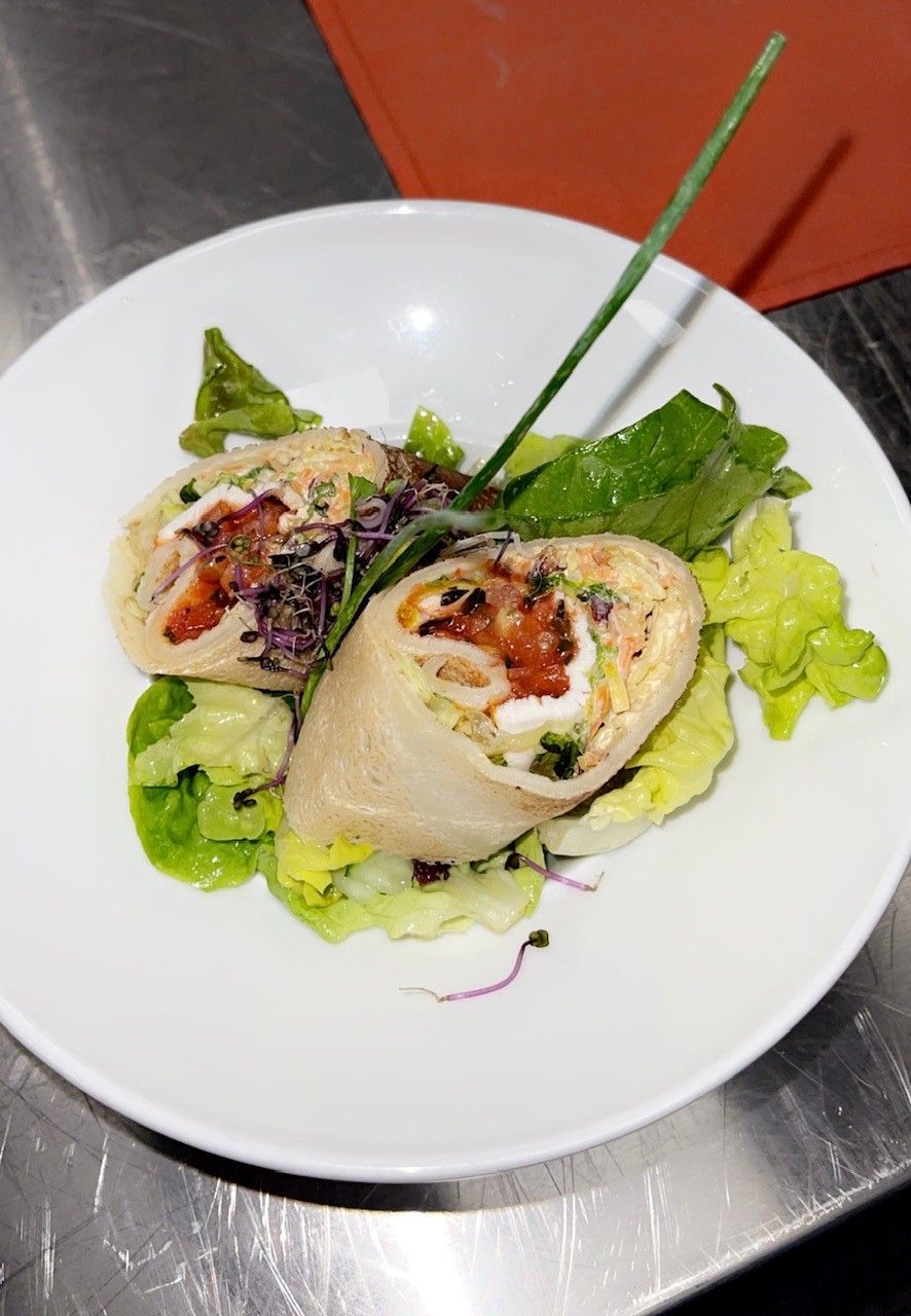 Prato com um alimento embalado, possivelmente um burrito, decorado com folhas verdes e molho, sobre um prato branco com alface embaixo.