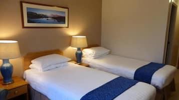 Quarto de hotel com duas camas, cada uma com lençóis brancos e um cobertor azul. Há duas luminárias de cabeceira, uma em cada cama. Um quadro emoldurado está pendurado na parede acima das camas.