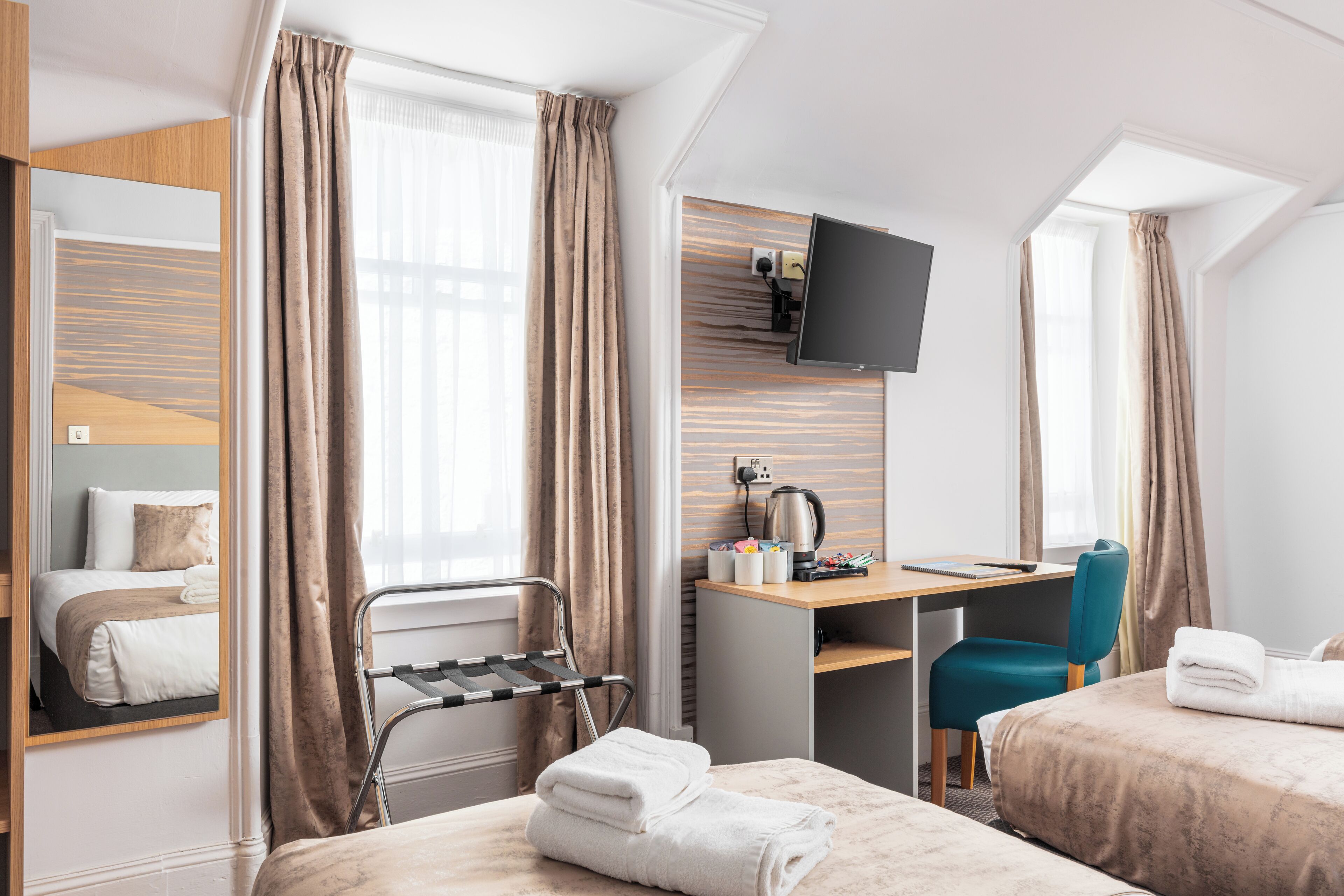 Quarto de hotel com cama, escrivaninha, cadeira, televisão e espelho.