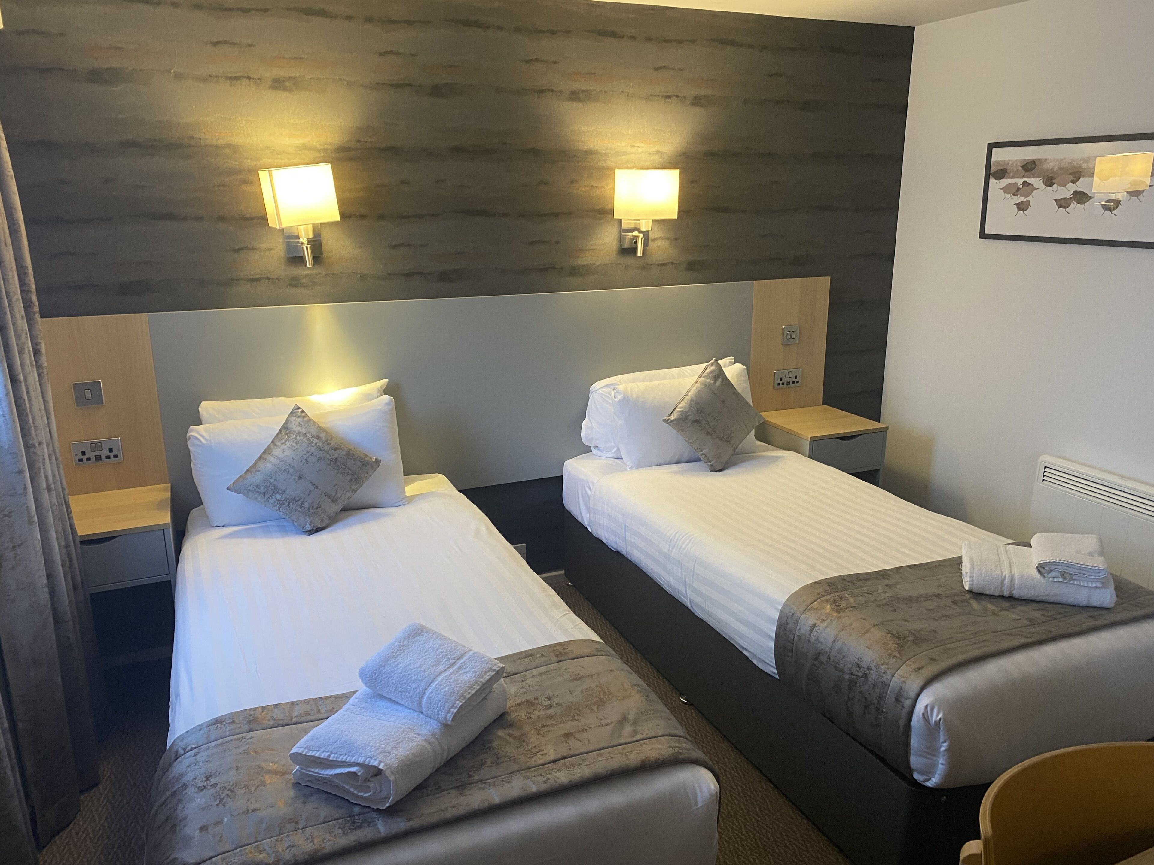 Quarto de hotel com duas camas, cada uma com lençóis e almofadas brancas, cabeceira de madeira, abajures, um quadro na parede e uma cadeira.