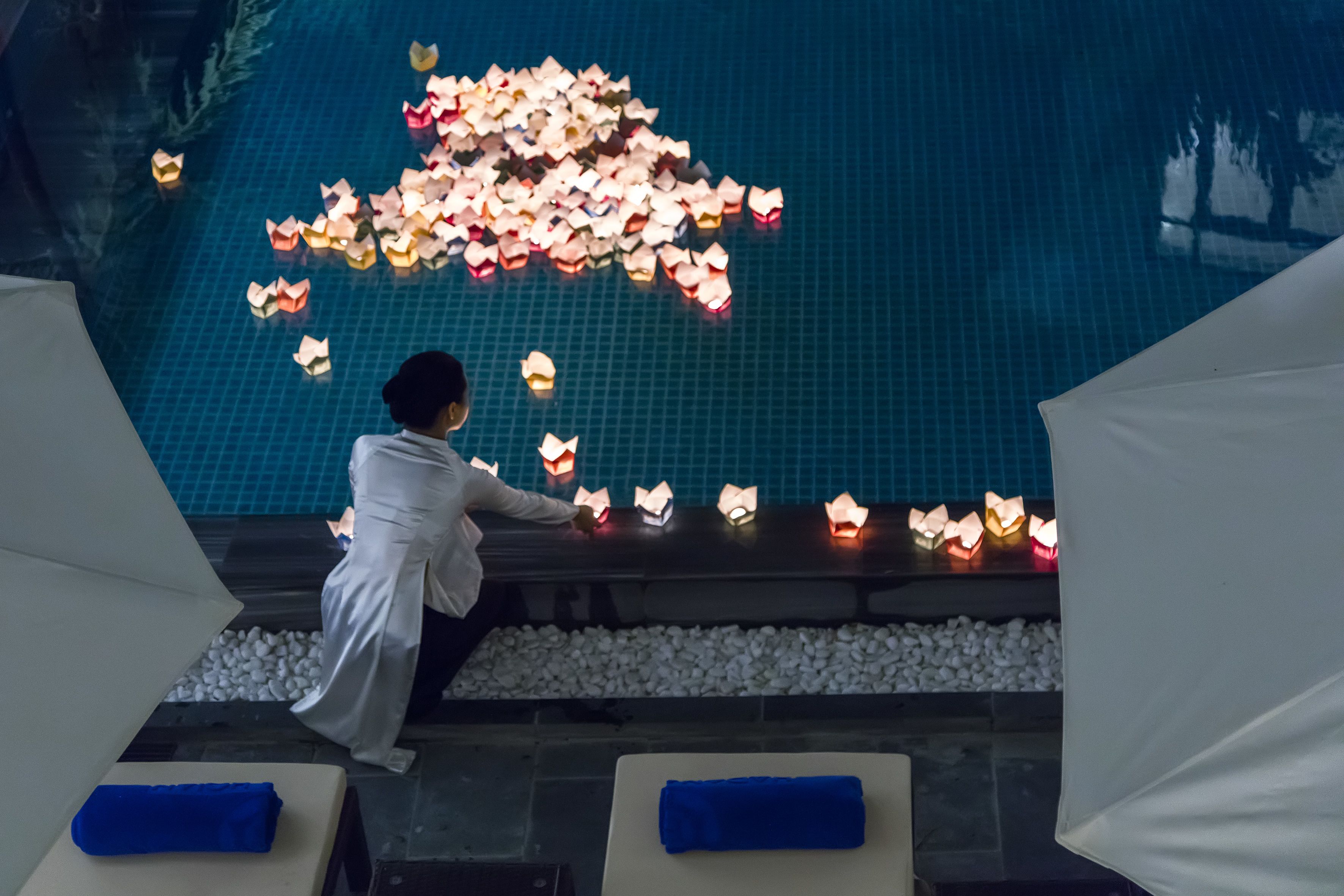 Uma área à beira da piscina com piso de azulejos, uma pessoa organizando velas flutuantes e cadeiras de descanso com almofadas azuis.