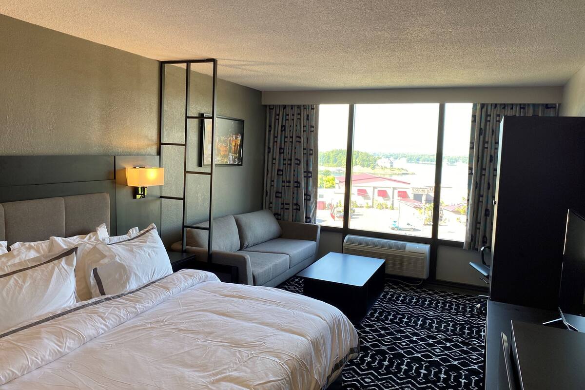 Quarto de hotel com cama, sofá, televisão, escrivaninha e uma grande janela com vista para prédios e áreas verdes.