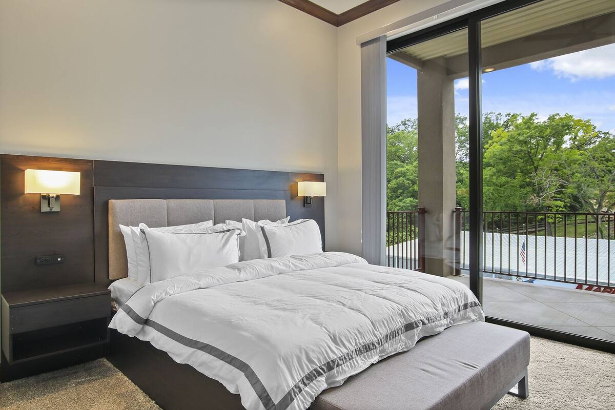 Quarto de hotel moderno com uma cama grande, duas luminárias de cabeceira, um banquinho e uma varanda com vista para árvores e uma bandeira.