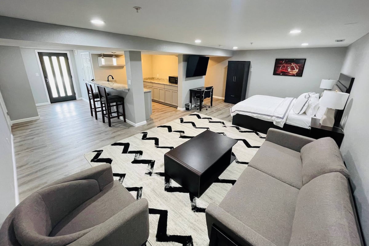 Quarto de hotel moderno com sofá, cama, TV e área de cozinha ao fundo.