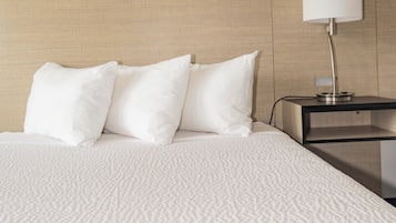 Uma cama bem arrumada com um espesso cobreleira branca e três travesseiros brancos. Uma mesa de cabeceira com abajur é visível à direita.