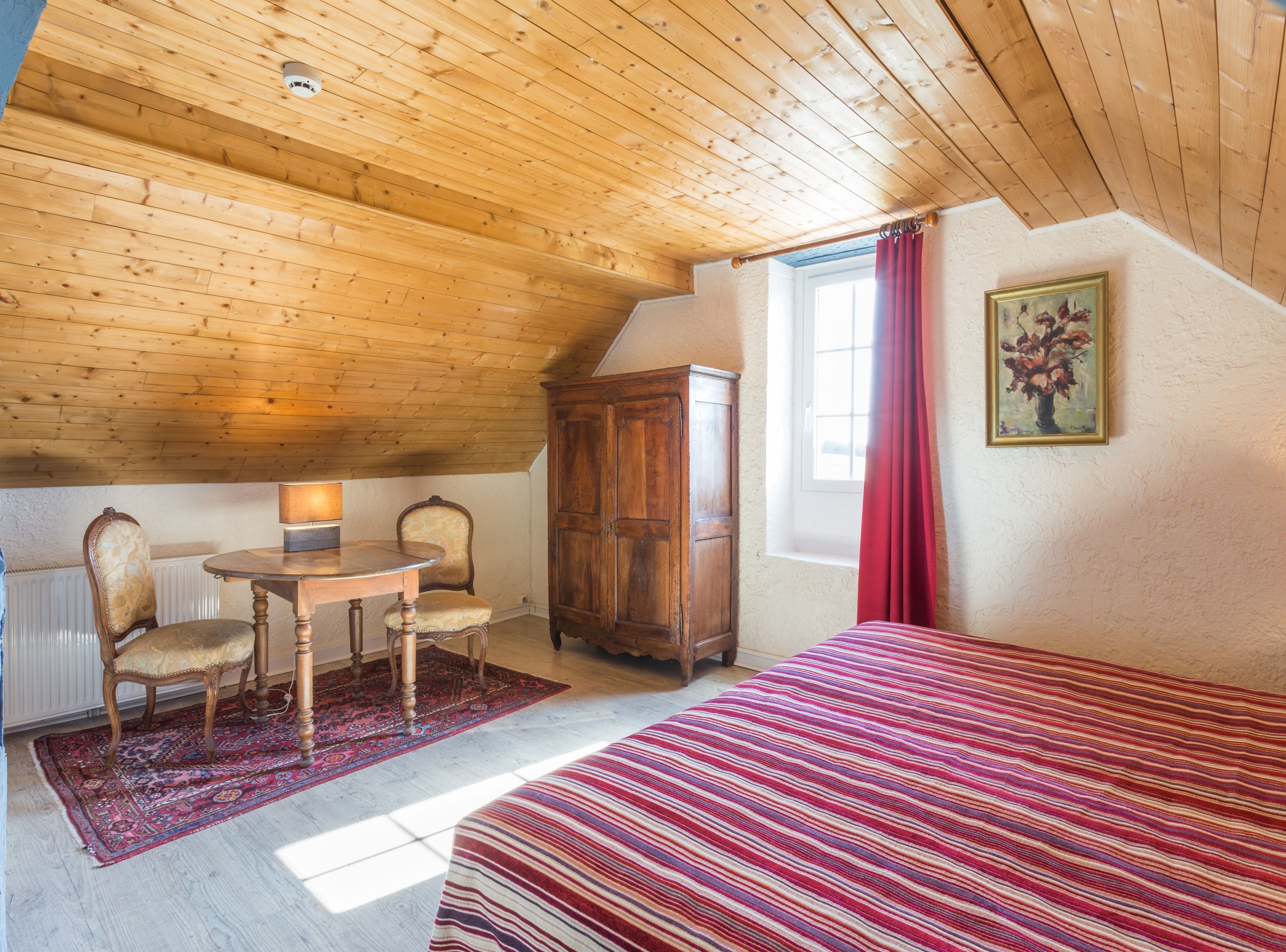 Um quarto de sótão aconchegante com teto de madeira, uma cama com colcha listrada, um armário de madeira, uma mesa com cadeiras e uma janela com cortinas vermelhas.