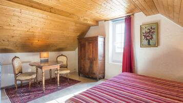 Um quarto de sótão aconchegante com teto de madeira, uma cama com colcha listrada, um armário de madeira, uma mesa com cadeiras e uma janela com cortinas vermelhas.
