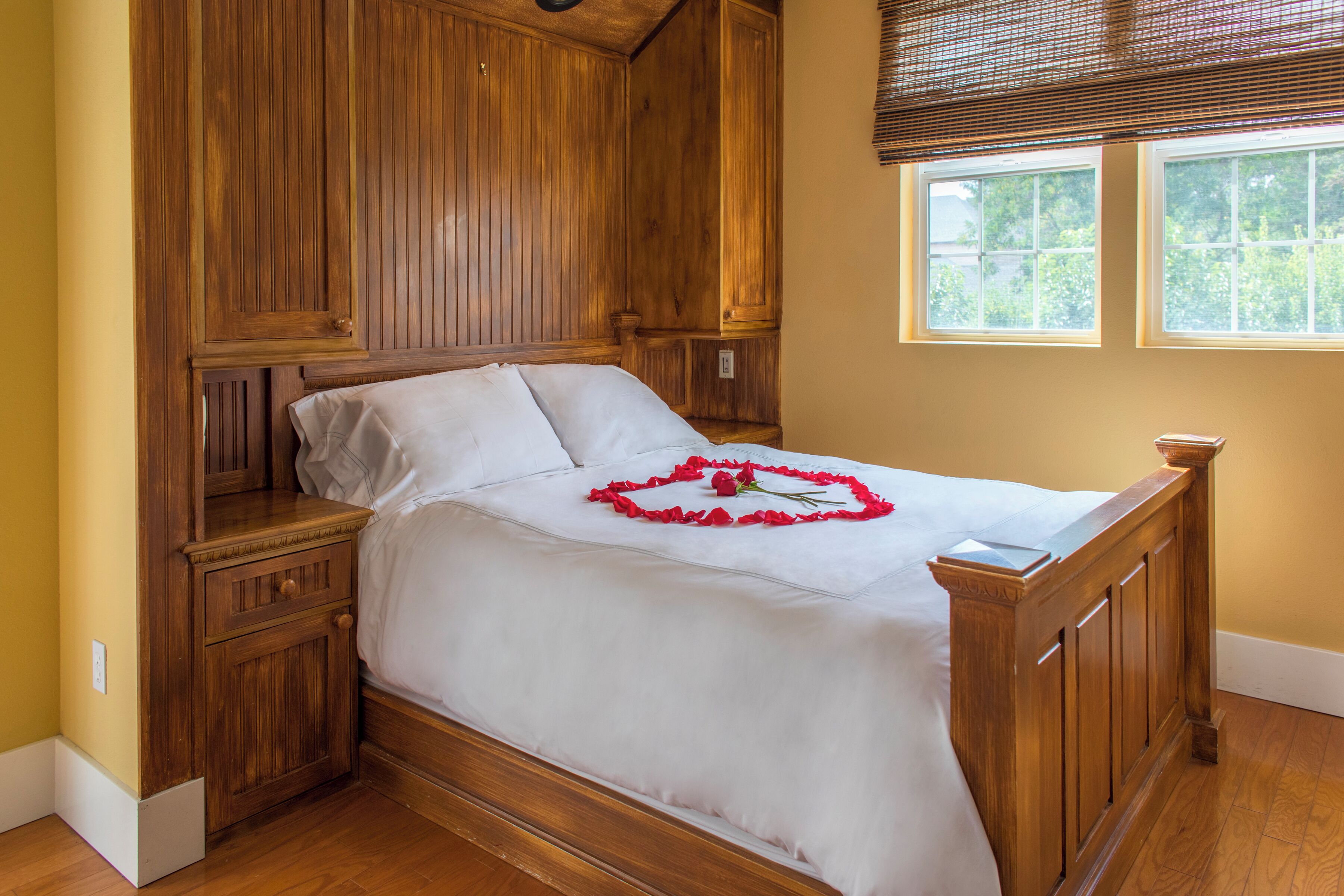 Uma cama de madeira com dossel, coberta por um edredom branco, e um arranjo de pétalas de rosas vermelhas em formato de coração.
