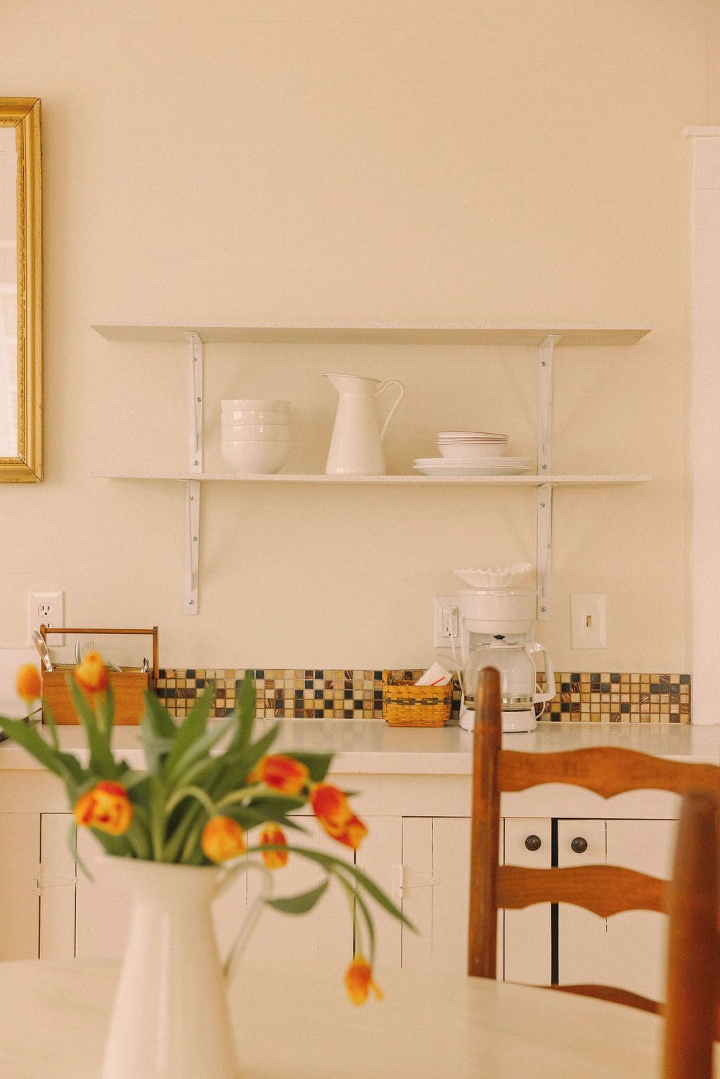 Uma cozinha com prateleiras brancas, uma cafeteira e um vaso com flores no balcão.