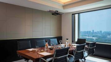 Sala de conferências com uma mesa grande, cadeiras pretas, projetor e vista para a cidade através de amplas janelas.