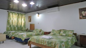 Quarto com três camas, cada uma com roupa de cama verde e padrão xadrez. A cama à esquerda tem uma cabeceira de madeira, a cama à direita possui uma mesa de cabeceira com abajur, e um ar-condicionado fixado na parede acima das camas.