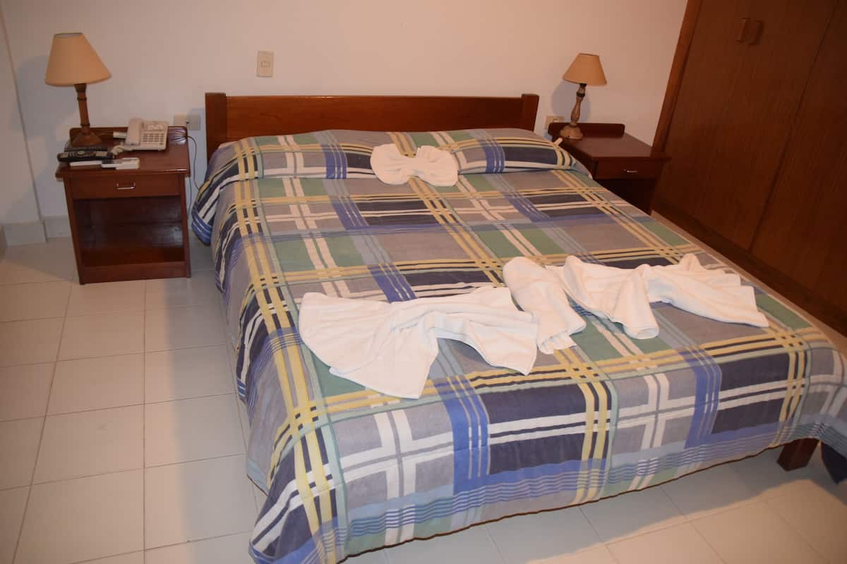 Uma cama bem arrumada com colcha xadrez, duas mesas de cabeceira com abajures, um telefone e uma gaveta pequena.
