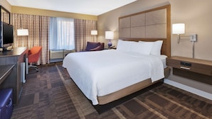 Quarto de hotel com uma cama grande, uma escrivaninha com televisão, uma cadeira e uma luminária de mesa.