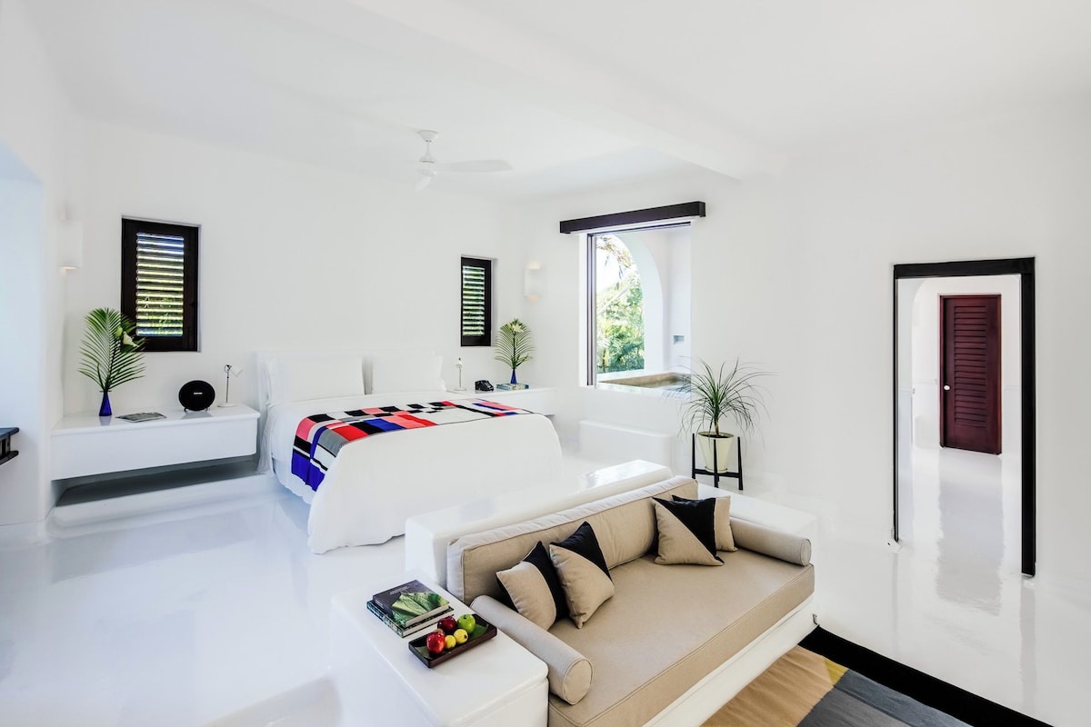 Quarto moderno com cama, sofá e uma pequena mesa com livros e frutas.