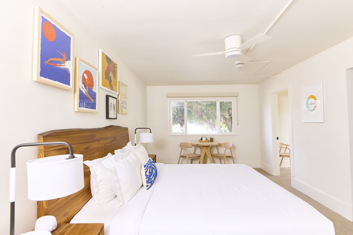 Quarto de hotel com uma cama grande, mesa de cabeceira com abajur, pequena mesa com cadeiras e obras de arte nas paredes.