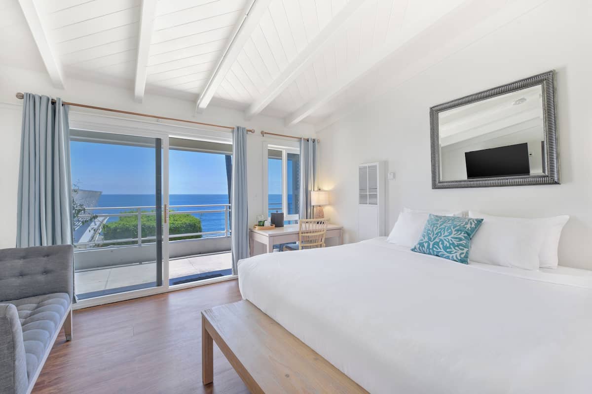 Quarto de hotel com uma cama grande, uma escrivaninha, uma cadeira e vista para o oceano através de uma porta de vidro deslizante.